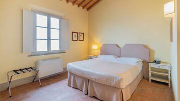 Quarto com cama, mesa de cabeceira, abajur, radiador e janela.