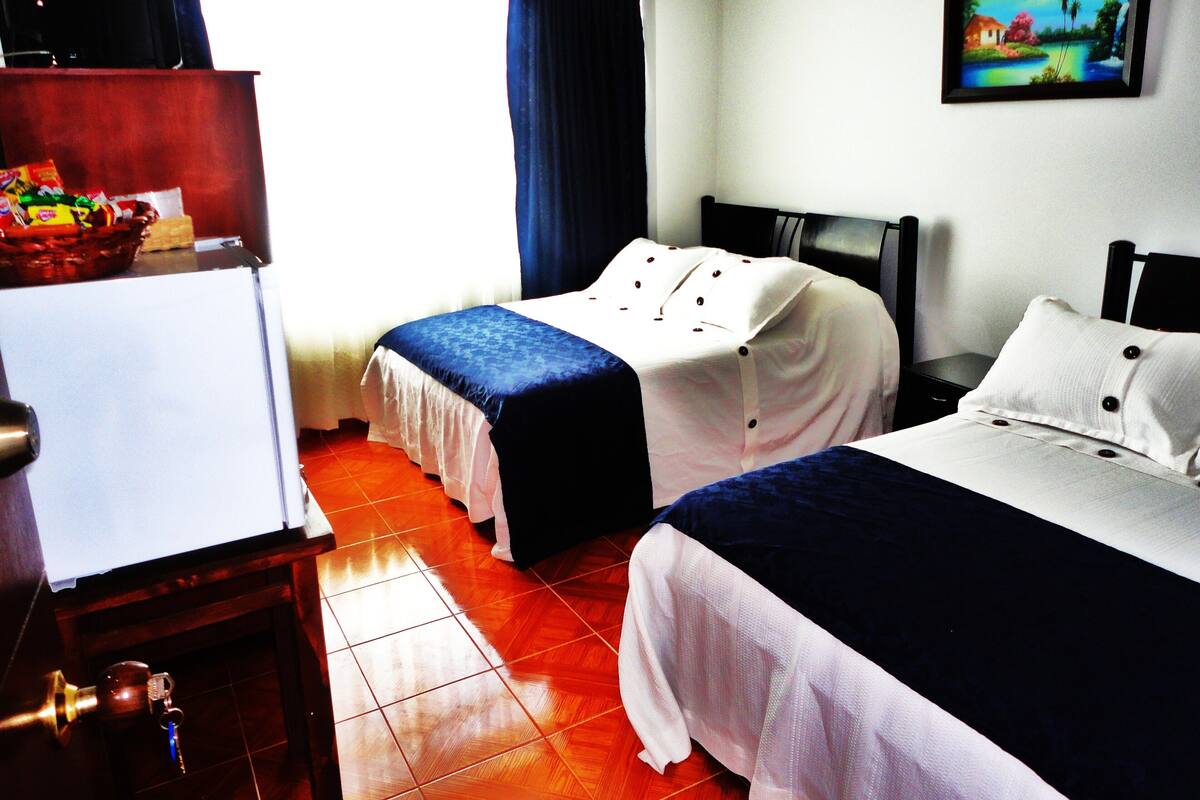 Quarto de hotel com duas camas, uma escrivaninha pequena, um frigorífico e um quadro na parede.