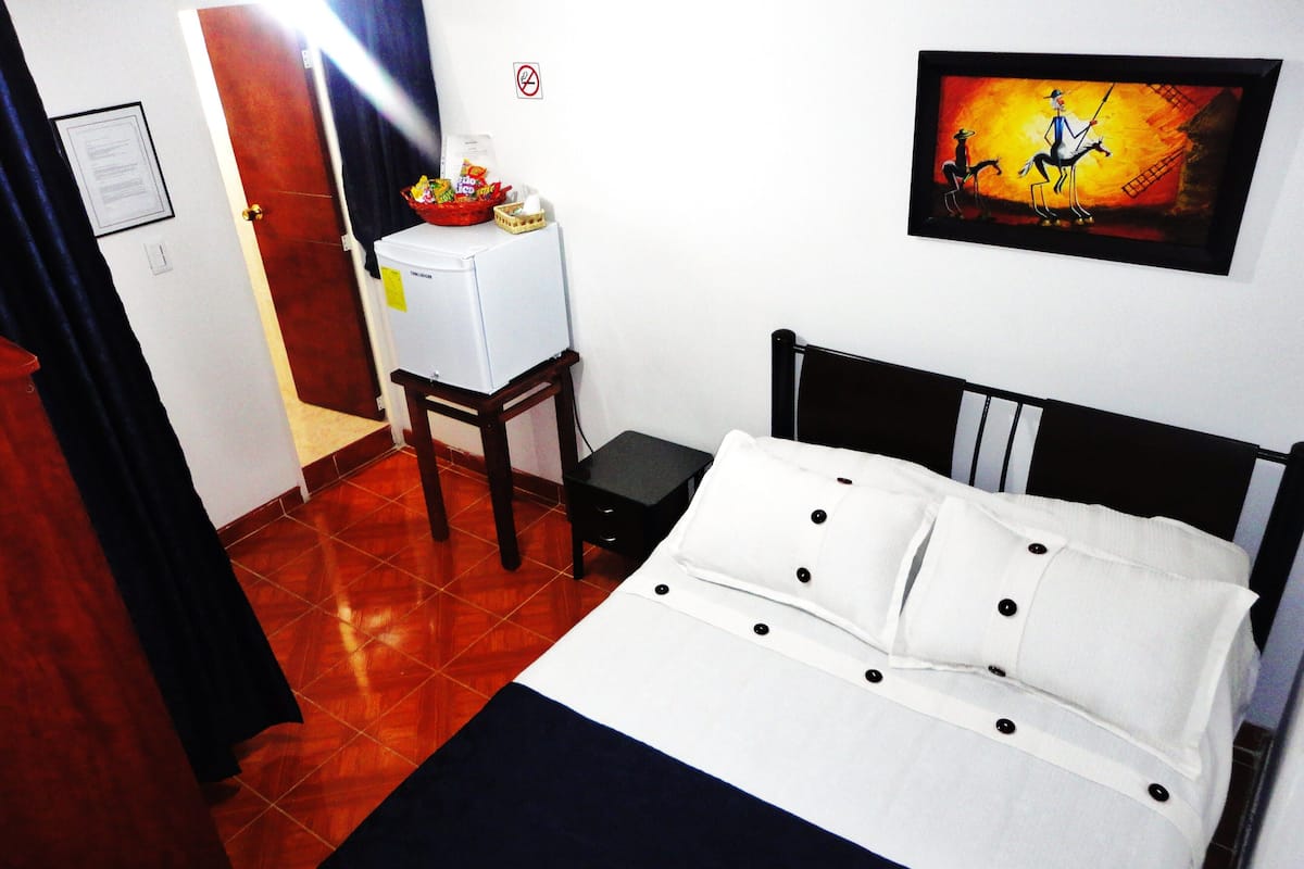 Quarto de hotel com uma cama, uma mesinha, um refrigerador e um quadro na parede.