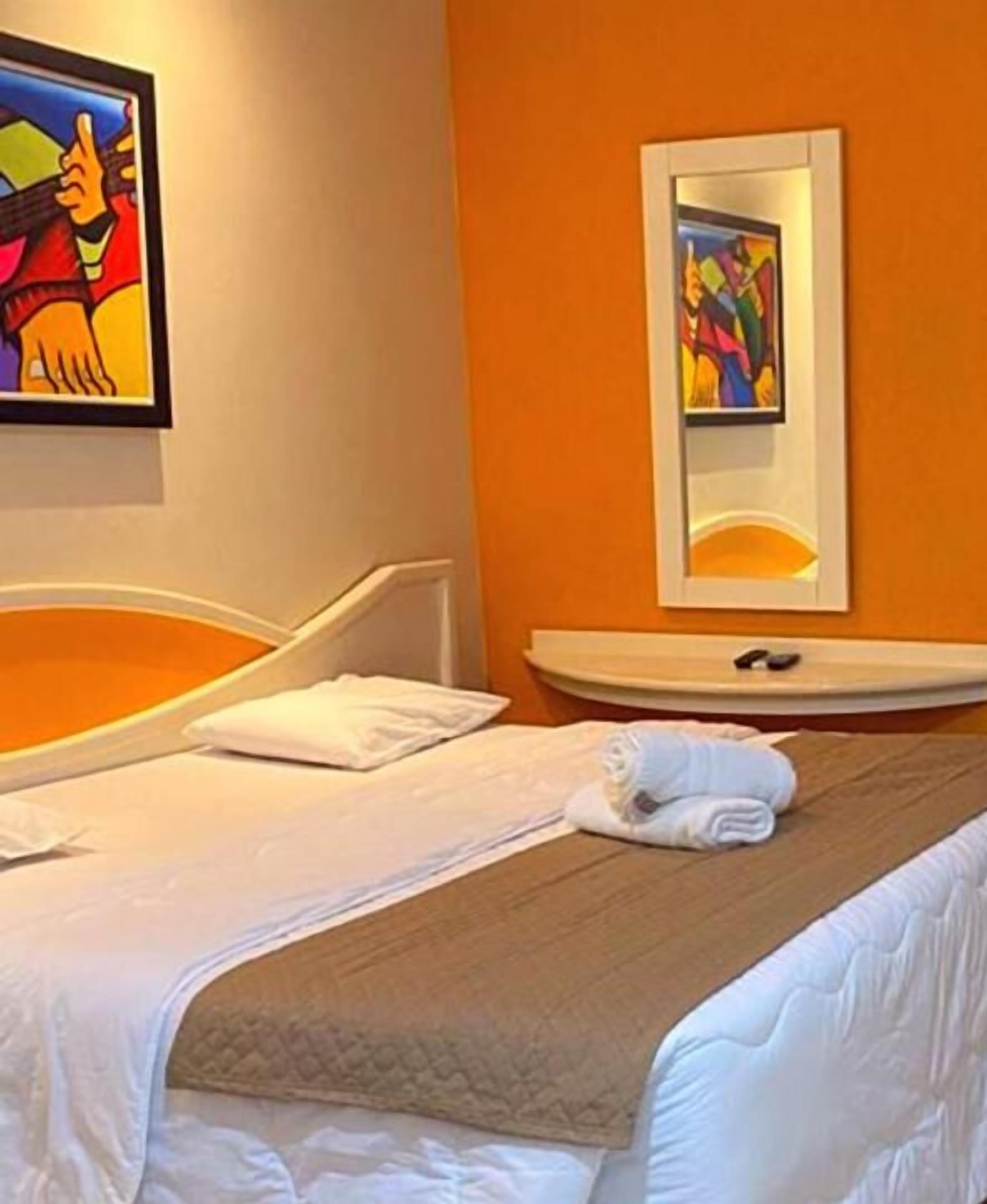 Quarto de hotel com uma cama, mesa de cabeceira, espelho e duas pinturas na parede.