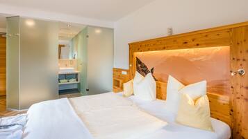 Quarto com cabeceira de madeira, cama com lençóis brancos e banheiro visível por uma porta aberta.