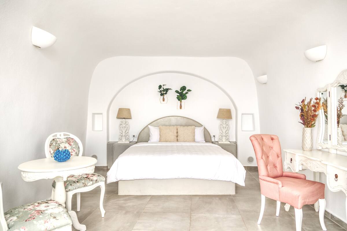 Quarto com uma cama grande, uma penteadeira com espelho, uma cadeira e uma mesinha com um vaso.