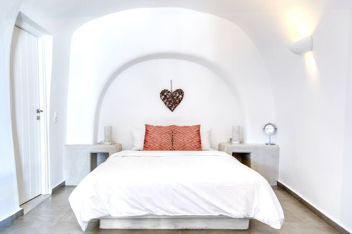 Quarto com cama branca, decoração em formato de coração na parede e duas mesas de cabeceira com abajures.
