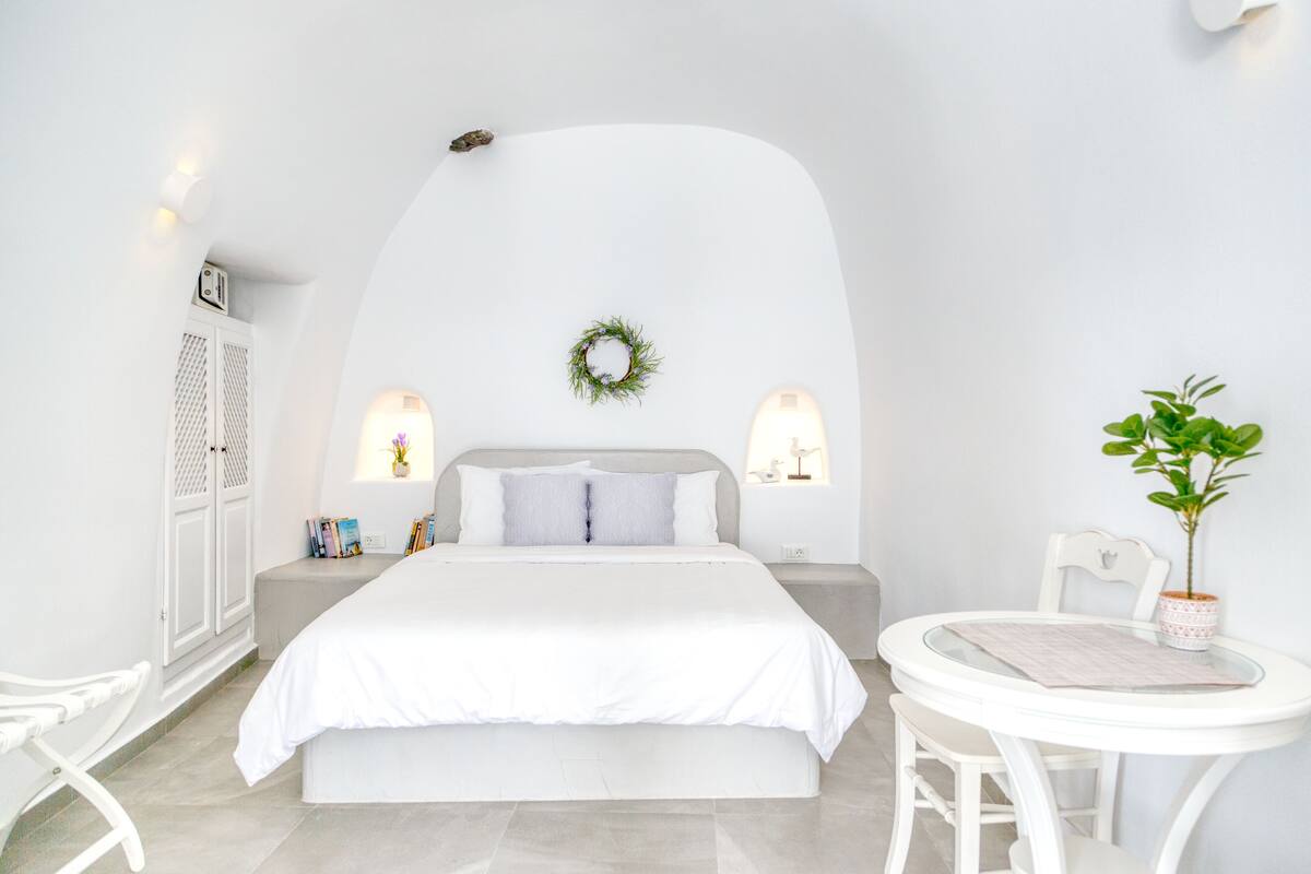 Quarto minimalista com cama, abajures, uma mesinha com uma planta e um luminário fixado na parede.