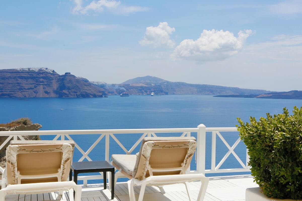 Terraço com duas cadeiras e uma mesa, com vista para um mar tranquilo e montanhas ao longe.