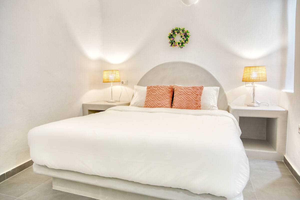 Cama bem arrumada com roupa de cama branca e duas almofadas decorativas, com mesas de cabeceira de cada lado com abajures.