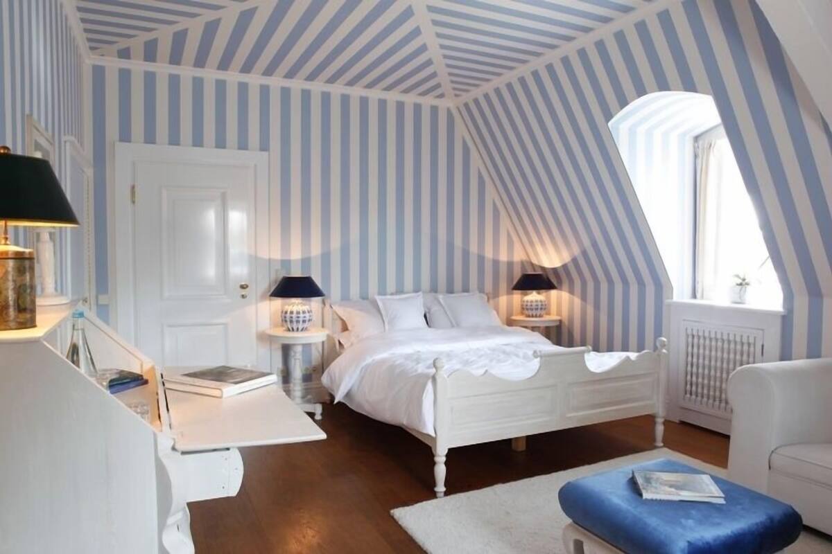 Quarto com cama branca, paredes listradas e teto inclinado. Há luminárias de cabeceira e uma janela com cortinas.
