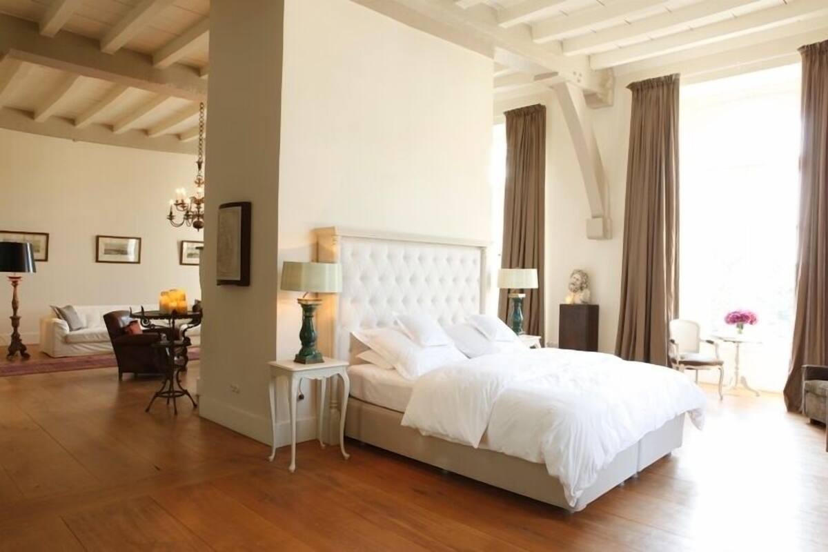 Quarto com uma cama grande, cabeceira branca, mesa de cabeceira com abajur, uma cadeira e uma pequena mesa com um vaso de flores.