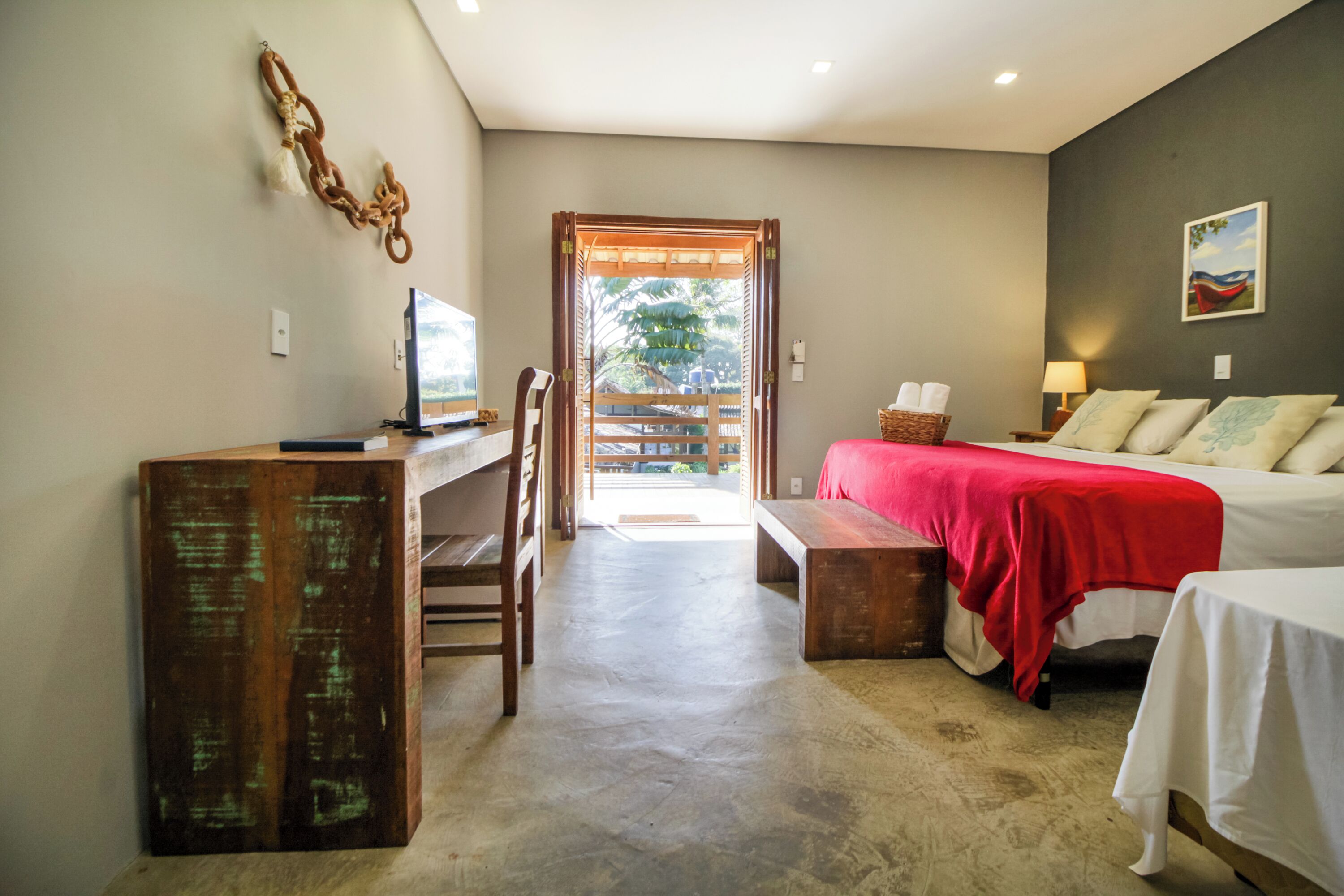 Quarto com cama, escrivaninha e computador. Tem uma janela com vista para árvores e uma porta que dá para o exterior.