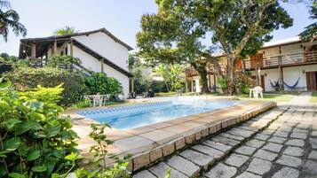 Uma piscina rodeada por azulejos de pedra, com um edifício branco e uma rede ao fundo.