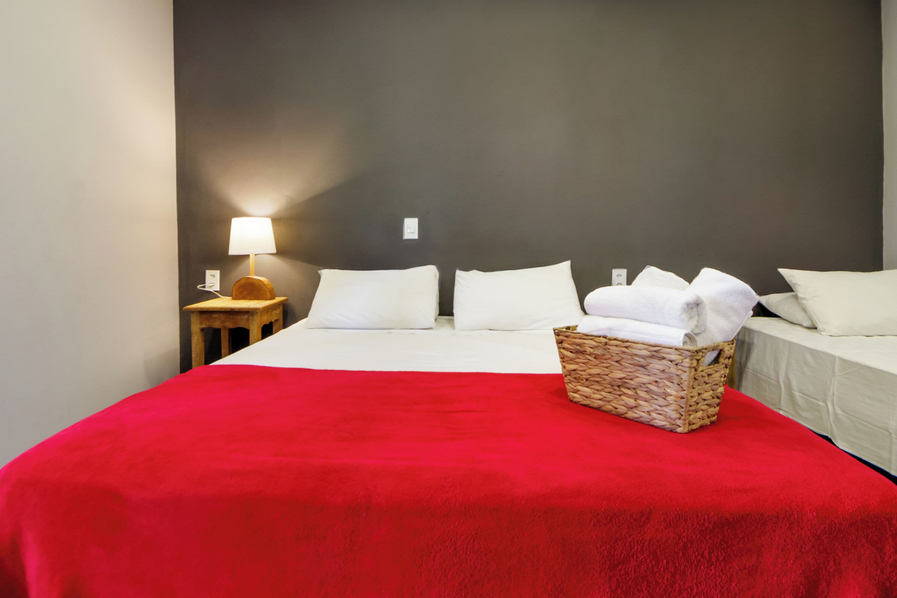 Quarto de hotel com uma cama grande, um cobertor vermelho, almofadas brancas, um abajur e um cesto de toalhas.