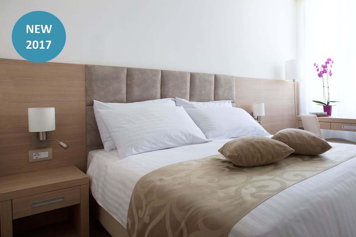 Uma cama bem arrumada com cabeceira bege, duas luminárias de cabeceira, um criado-mudo com gaveta e uma janela com cortinas.