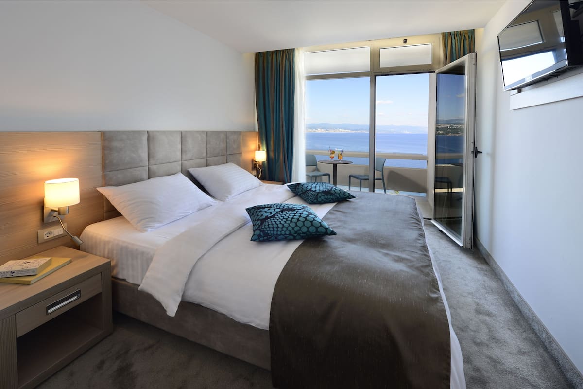Quarto de hotel com uma cama grande, mesa de cabeceira com luminária, vista para o mar através de uma varanda e uma mesa com cadeiras.