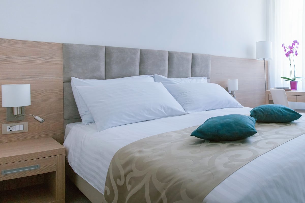 Cama bem arrumada com lençóis brancos e duas almofadas verde-água, mesa de cabeceira com abajur e apoio com tomada.