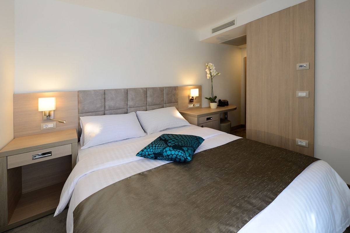 Quarto de hotel com cama, mesas de cabeceira, espelho e aquecedor fixado na parede.