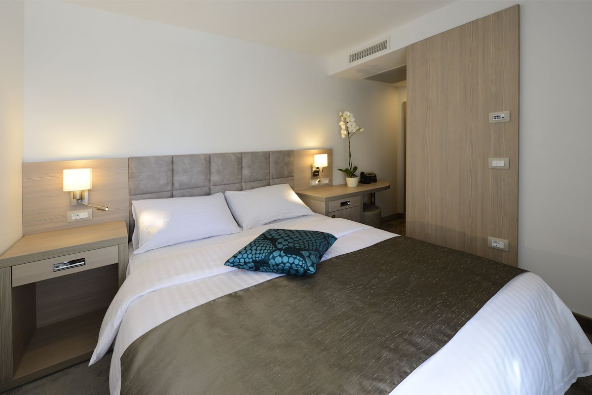 Quarto de hotel com cama, mesas de cabeceira, luminária e planta em vaso.