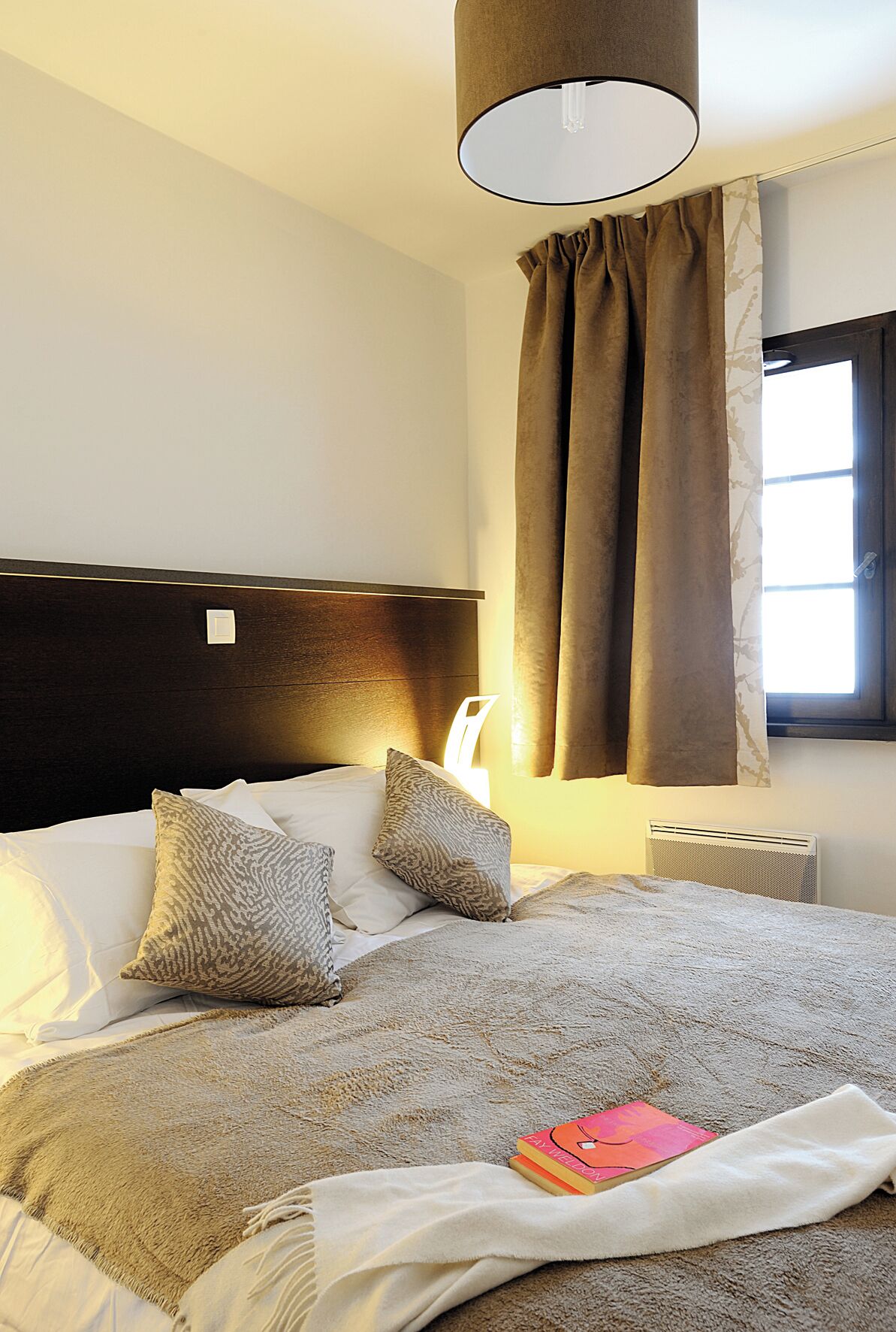 Quarto de hotel com cama, almofadas, mesa de cabeceira, luminária e janela com cortinas.