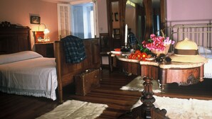 Quarto tradicional com cama, cómoda de madeira, espelho e mesa com flores e chapéu.