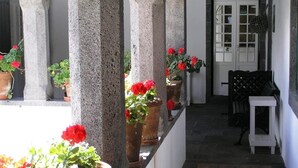 Área externa coberta com colunas de pedra, plantas em vasos, um banco e uma porta de entrada.