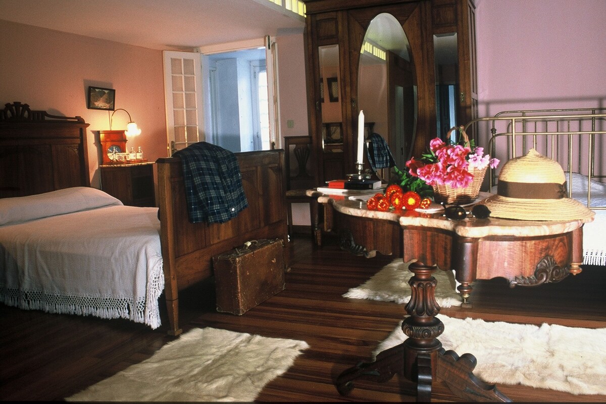 Quarto tradicional com cama, cómoda de madeira, espelho e mesa com flores e chapéu.