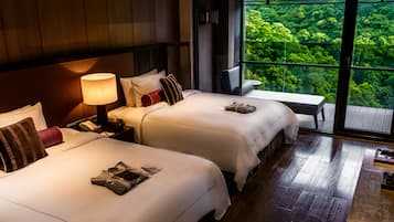 Quarto de hotel com duas camas, uma mesa de cabeceira, um abajur e vista para uma floresta através de grandes janelas.