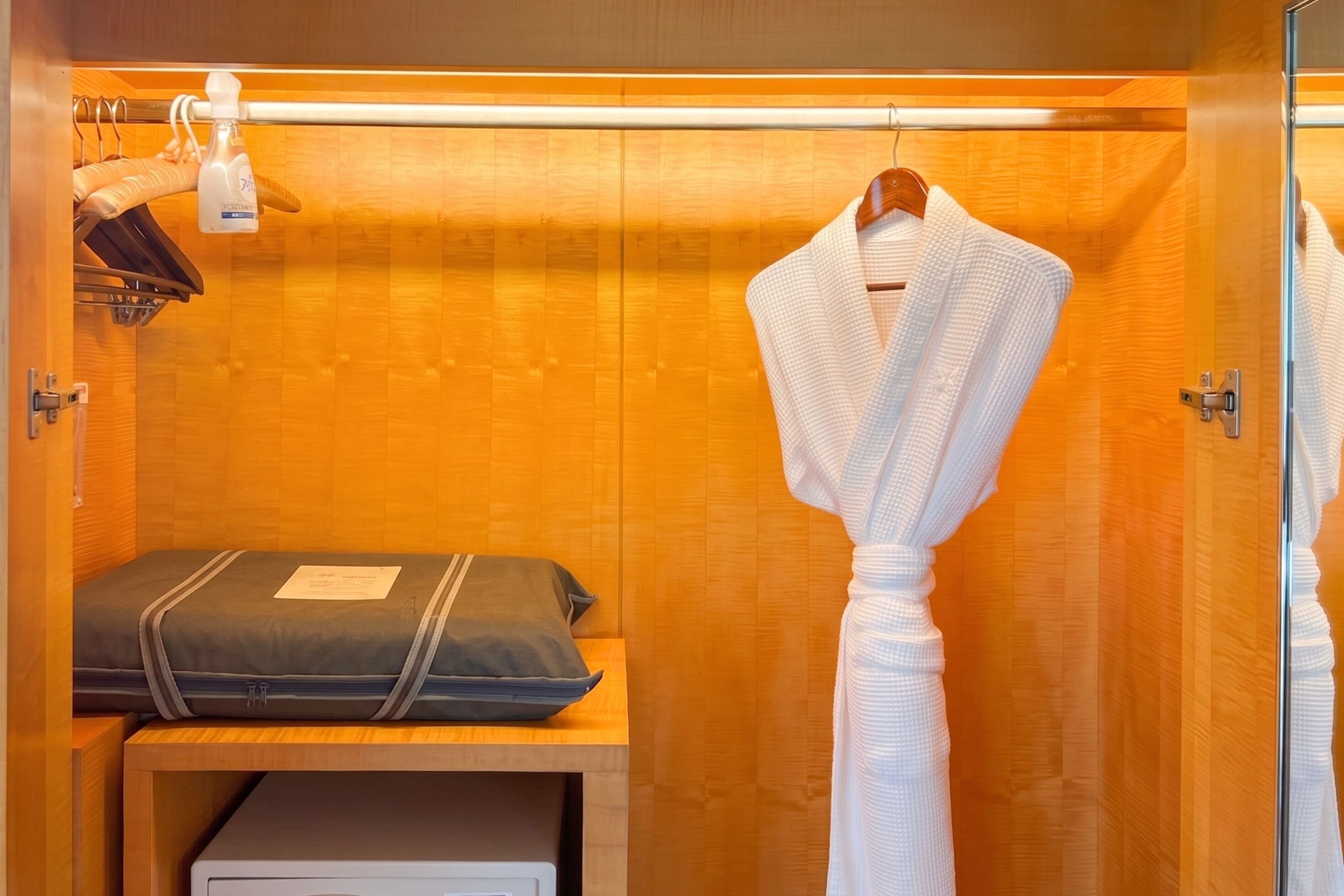 Quarto de hotel com parede de madeira, roupão pendurado em um gancho, porta-toalhas e uma prateleira com travesseiro e caixa.