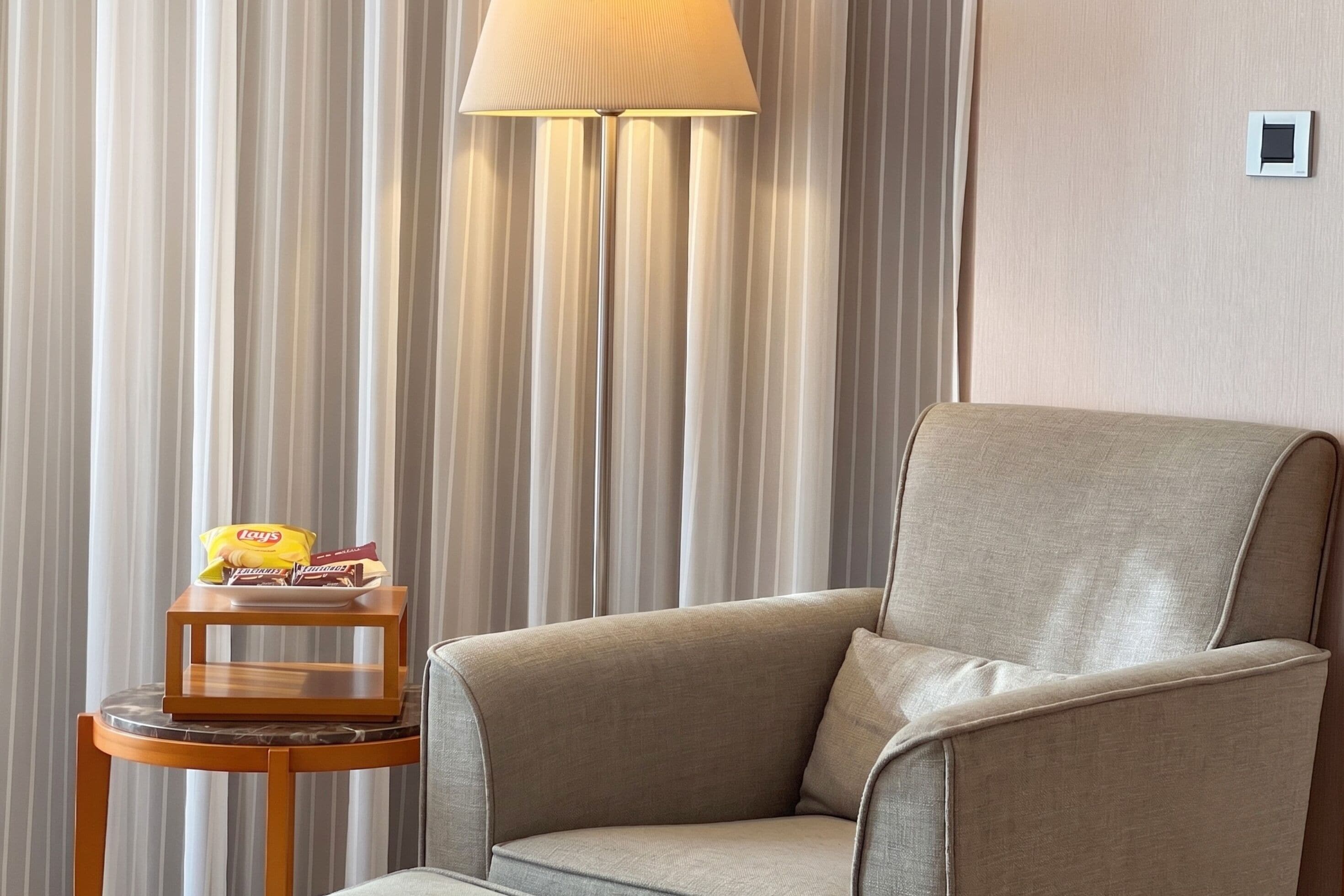Quarto de hotel com uma poltrona cinza, uma pequena mesa de cabeceira com lanches e uma luminária.
