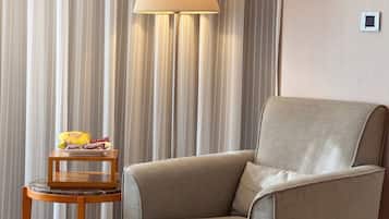 Quarto de hotel com uma poltrona cinza, uma pequena mesa de cabeceira com lanches e uma luminária.