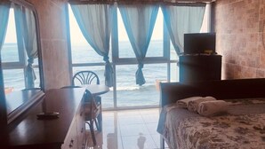 Quarto com cama, televisão, escrivaninha e vista para o oceano através de grandes janelas.