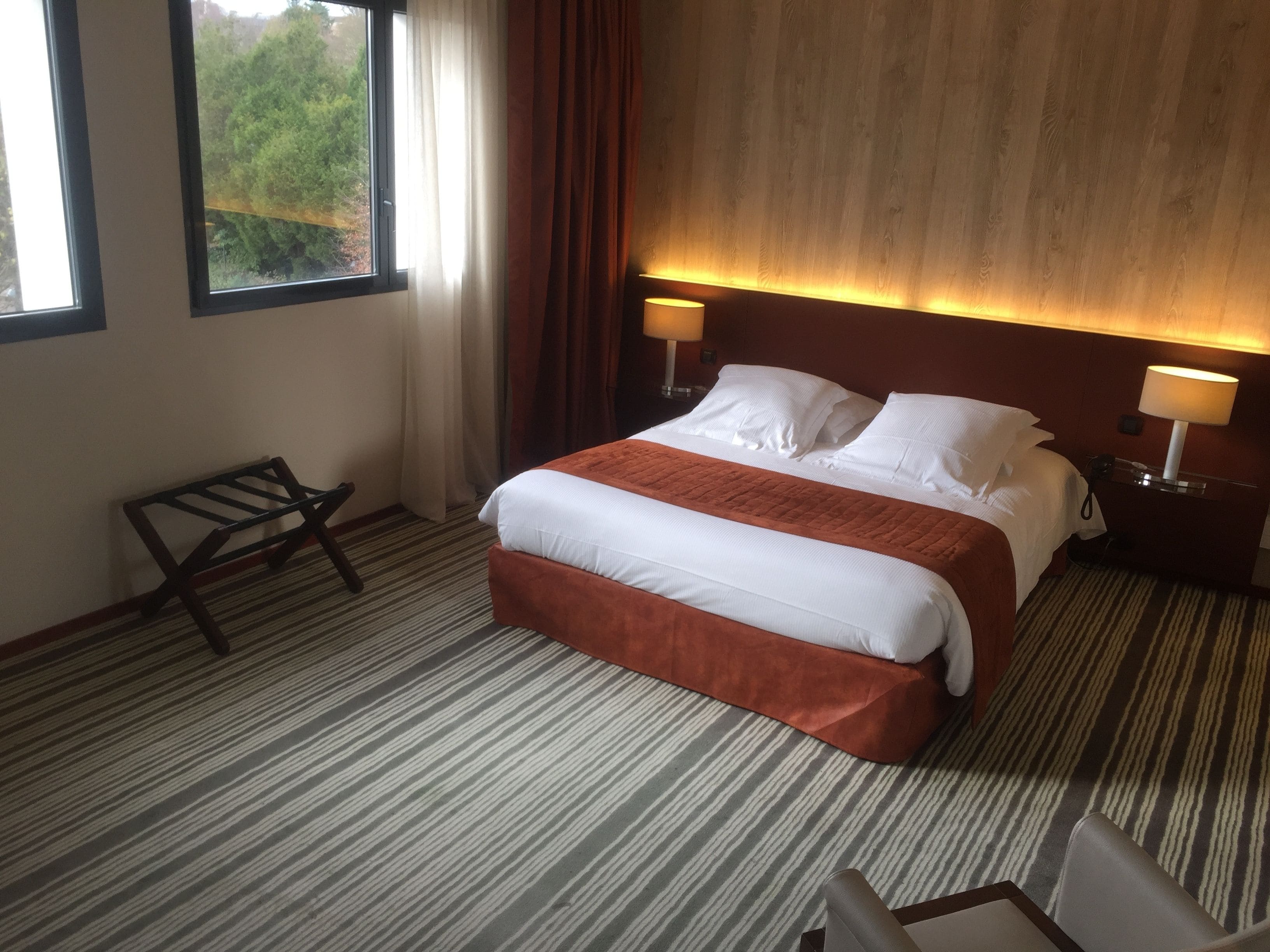 Quarto de hotel com uma cama grande, duas luminárias de cabeceira, um painel de cabeceira de madeira, um tapete listrado e uma janela com cortinas.