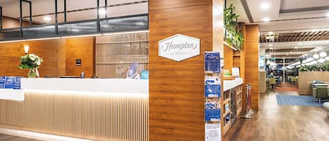 Uma área de recepção de hotel com balcão de madeira, uma placa com a inscrição "Hampton by Hilton" e um espaço de estar com mesas e cadeiras.