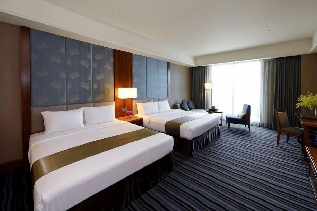 Quarto de hotel com duas camas, uma escrivaninha, uma cadeira e uma janela grande.