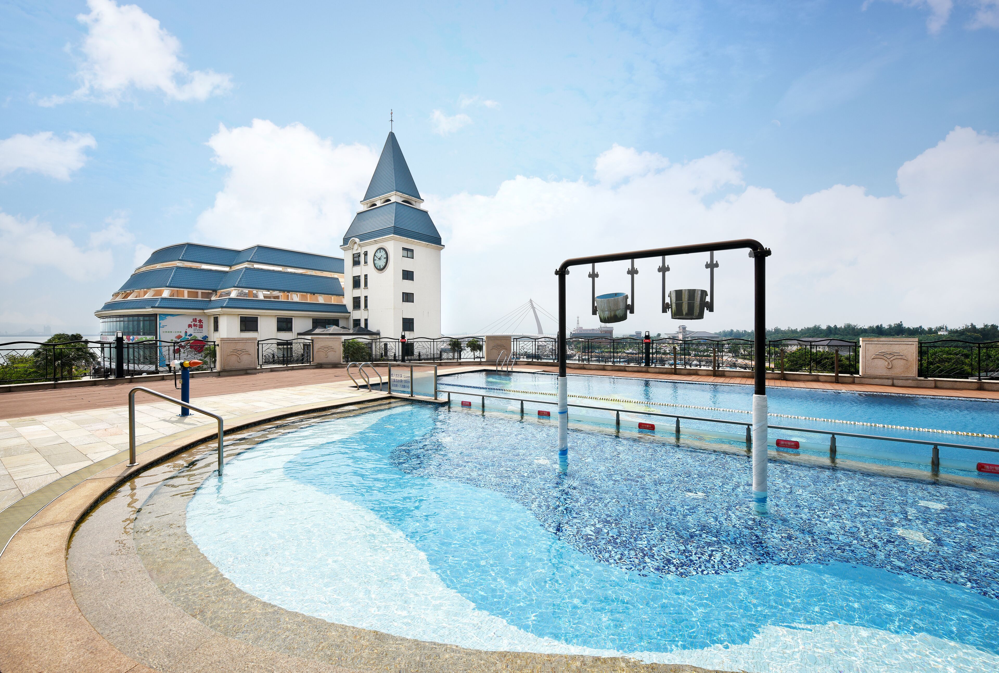 Piscina com uma torre do relógio ao fundo, rodeada por um terraço com guardaílos e vista para o horizonte da cidade.
