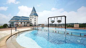 Piscina com uma torre do relógio ao fundo, rodeada por um terraço com guardaílos e vista para o horizonte da cidade.