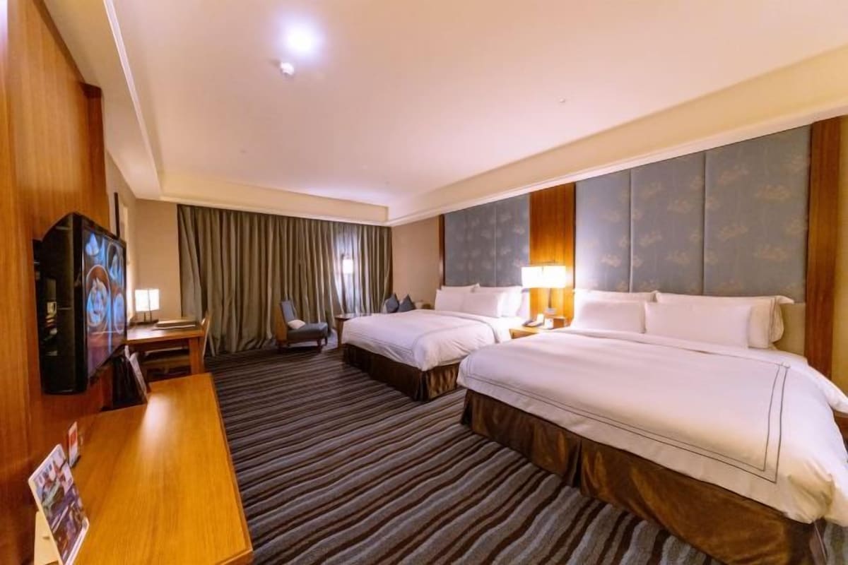 Quarto de hotel com duas camas, uma escrivaninha, uma cadeira, televisão e uma janela grande com cortinas.