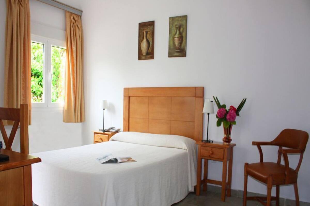Quarto de hotel com cama, mesa de cabeceira de madeira, uma cadeira, abajur e duas pinturas na parede.