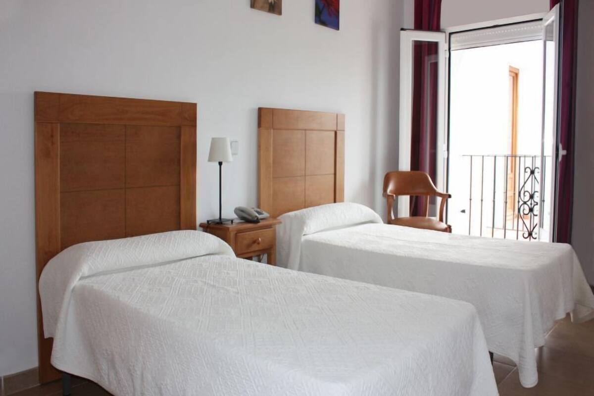 Quarto de hotel com duas camas de solteiro, mesa de cabeceira com abajur e telefone, painel de cama de madeira e vista para a varanda.