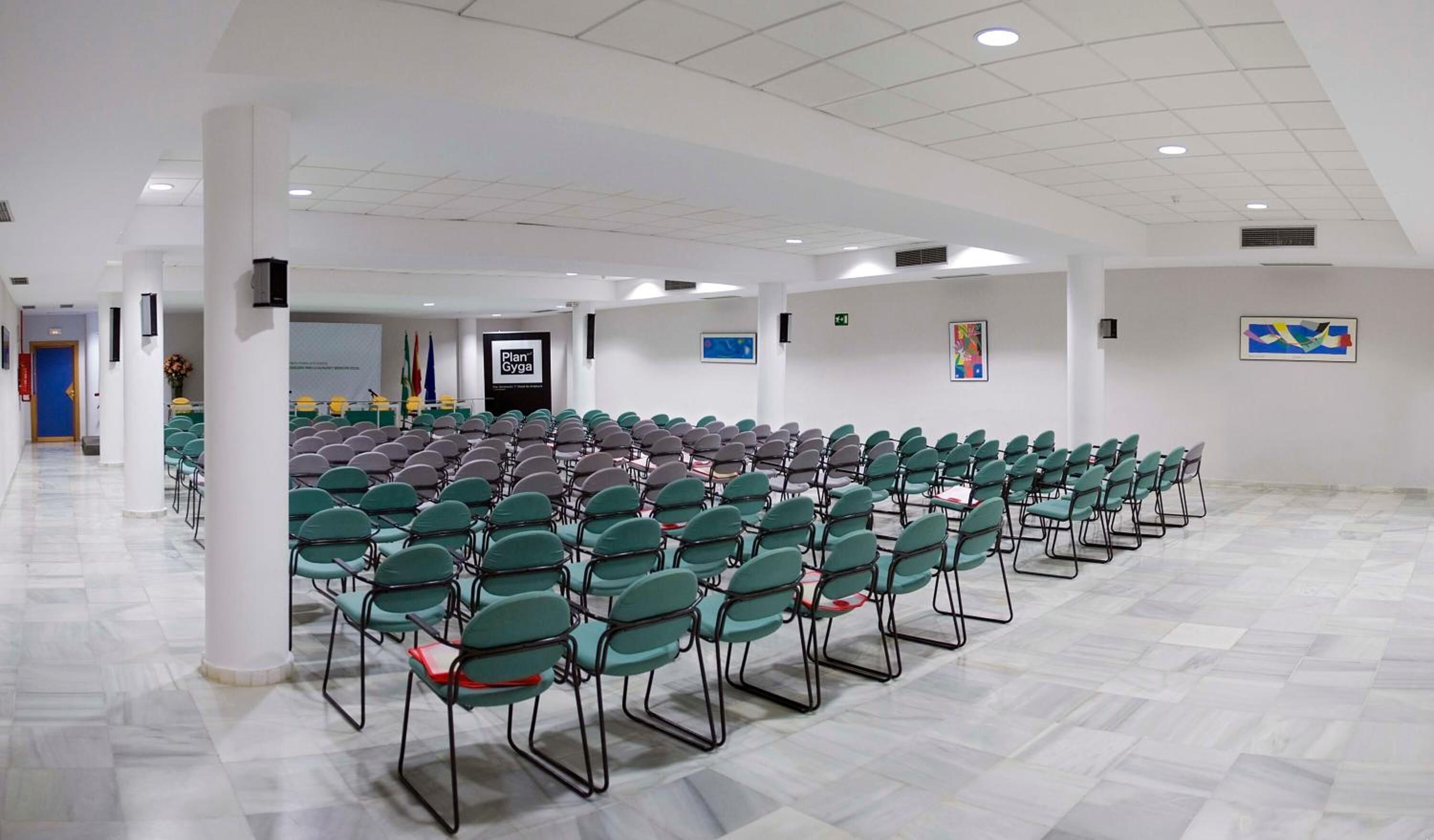 Uma sala de conferências com fileiras de cadeiras verdes, paredes brancas e chão de azulejos.