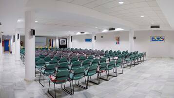 Uma sala de conferências com fileiras de cadeiras verdes, paredes brancas e chão de azulejos.