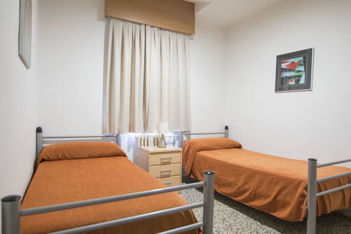 Quarto com duas camas de solteiro, uma mesinha com abajur, janela com cortinas e um quadro na parede.