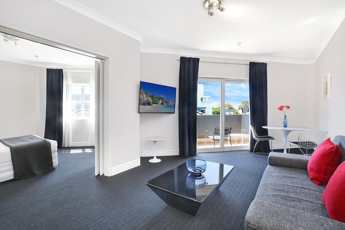 Quarto de hotel moderno com cama, mesa de centro de vidro, sofá e varanda com vista para o mar.