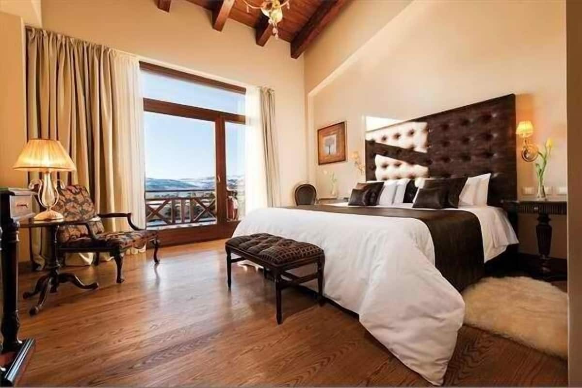 Quarto de hotel com uma cama grande, piso de madeira, vista para o exterior, uma escrivaninha com abajur e uma cadeira.