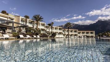 Um hotel com piscina, palmeiras e montanhas ao fundo.