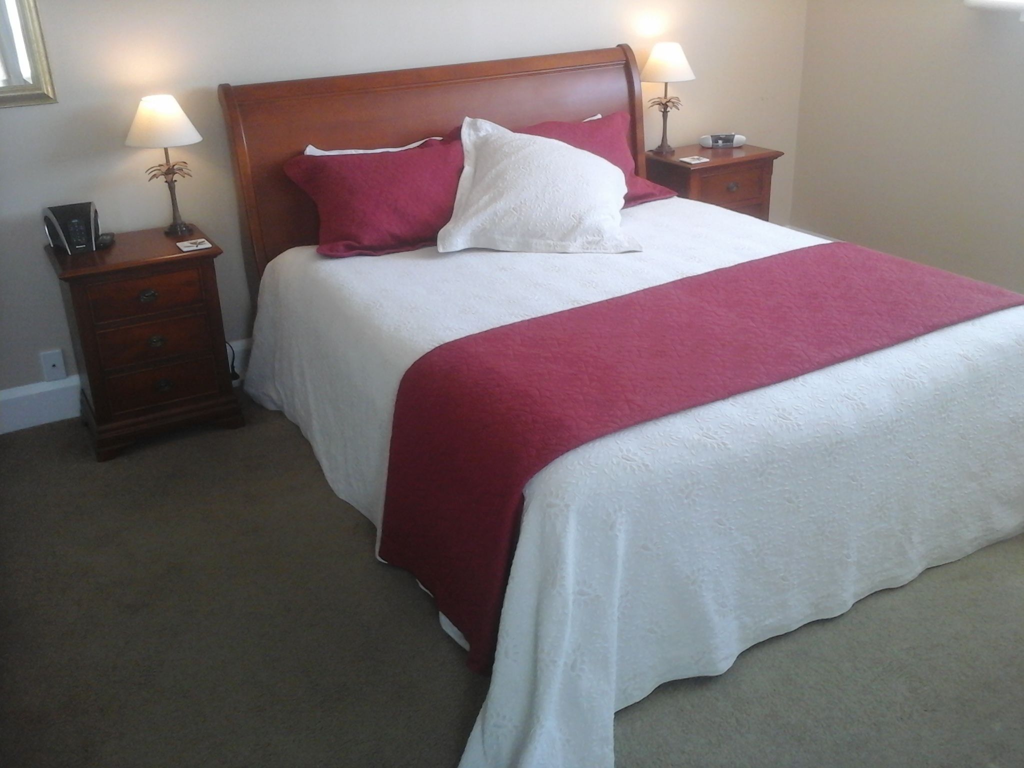 Cama bem arrumada com cabeceira de madeira, duas mesas de cabeceira com luminárias e um pequeno aparador de madeira.