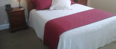 Cama bem arrumada com cabeceira de madeira, duas mesas de cabeceira com luminárias e um pequeno aparador de madeira.