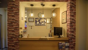 Uma área de recepção com balcão de madeira, parede de pedra, luminárias pendentes, um relógio, certificados emoldurados e uma placa indicando para deixar as chaves na recepção ao sair.