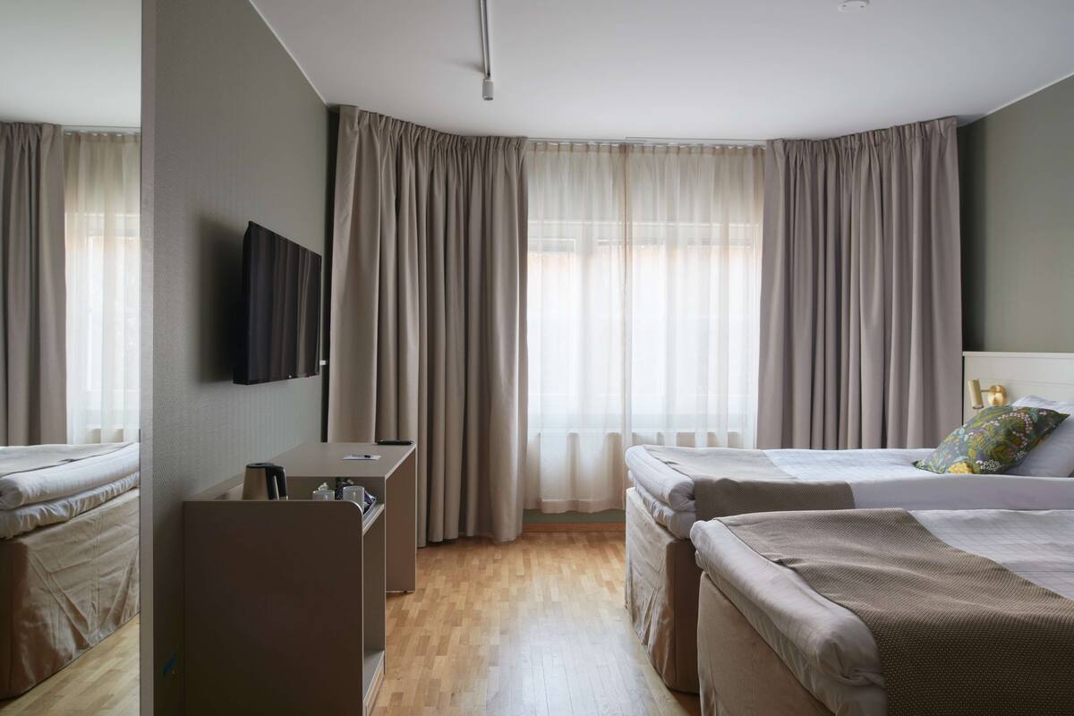 Quarto de hotel com duas camas, uma escrivaninha, televisão e uma janela grande com cortinas.