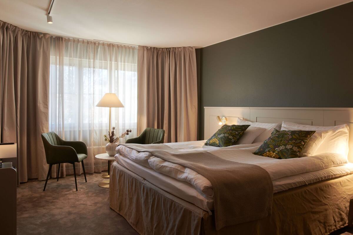 Quarto de hotel com uma cama grande, um espaço de estar com uma poltrona e uma mesinha, e uma janela com cortinas finas.
