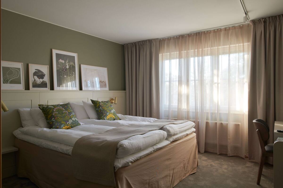 Cama bem arrumada com cabeceira, mesa de cabeceira e uma cadeira. Quadros emoldurados na parede acima da cama.