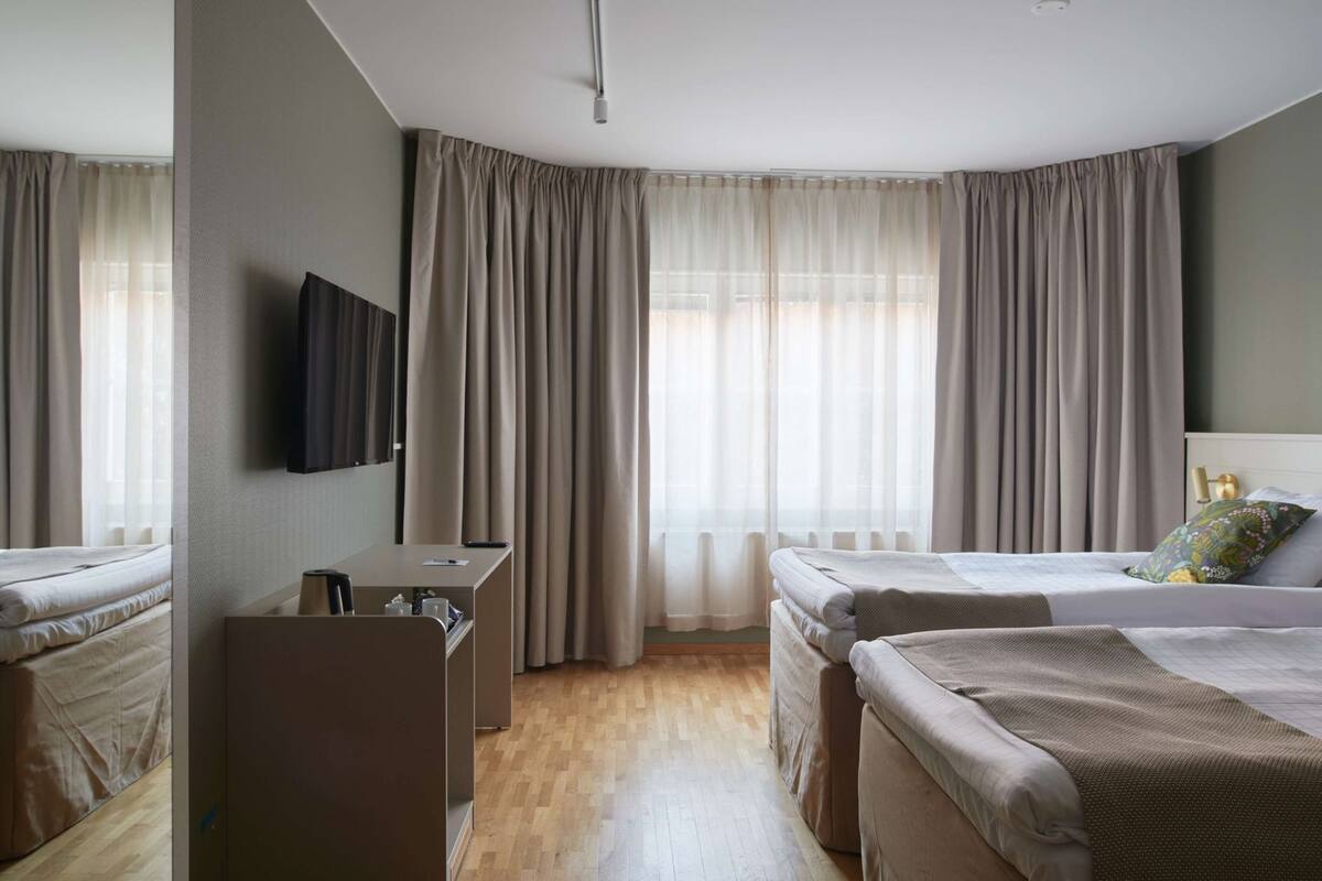 Quarto de hotel com duas camas, uma escrivaninha, televisão e janelas amplas com cortinas.